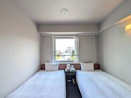 Quarto de hotel com duas camas, uma janela com vista para edifícios, uma mesa de cabeceira com luminária e uma mesinha com telefone e um copo.