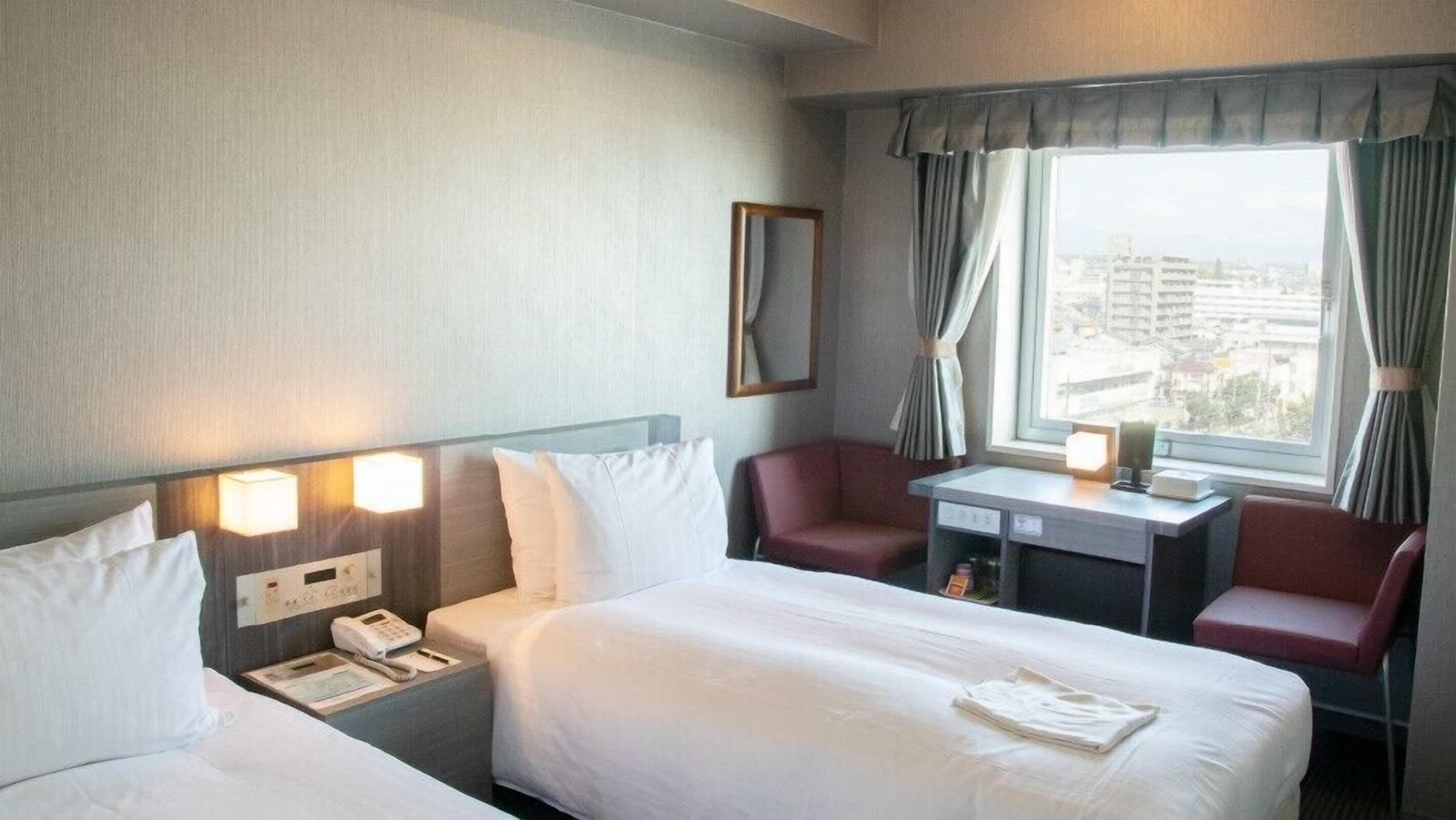 Quarto de hotel com duas camas, uma escrivaninha, uma cadeira, um telefone e uma janela com cortinas.
