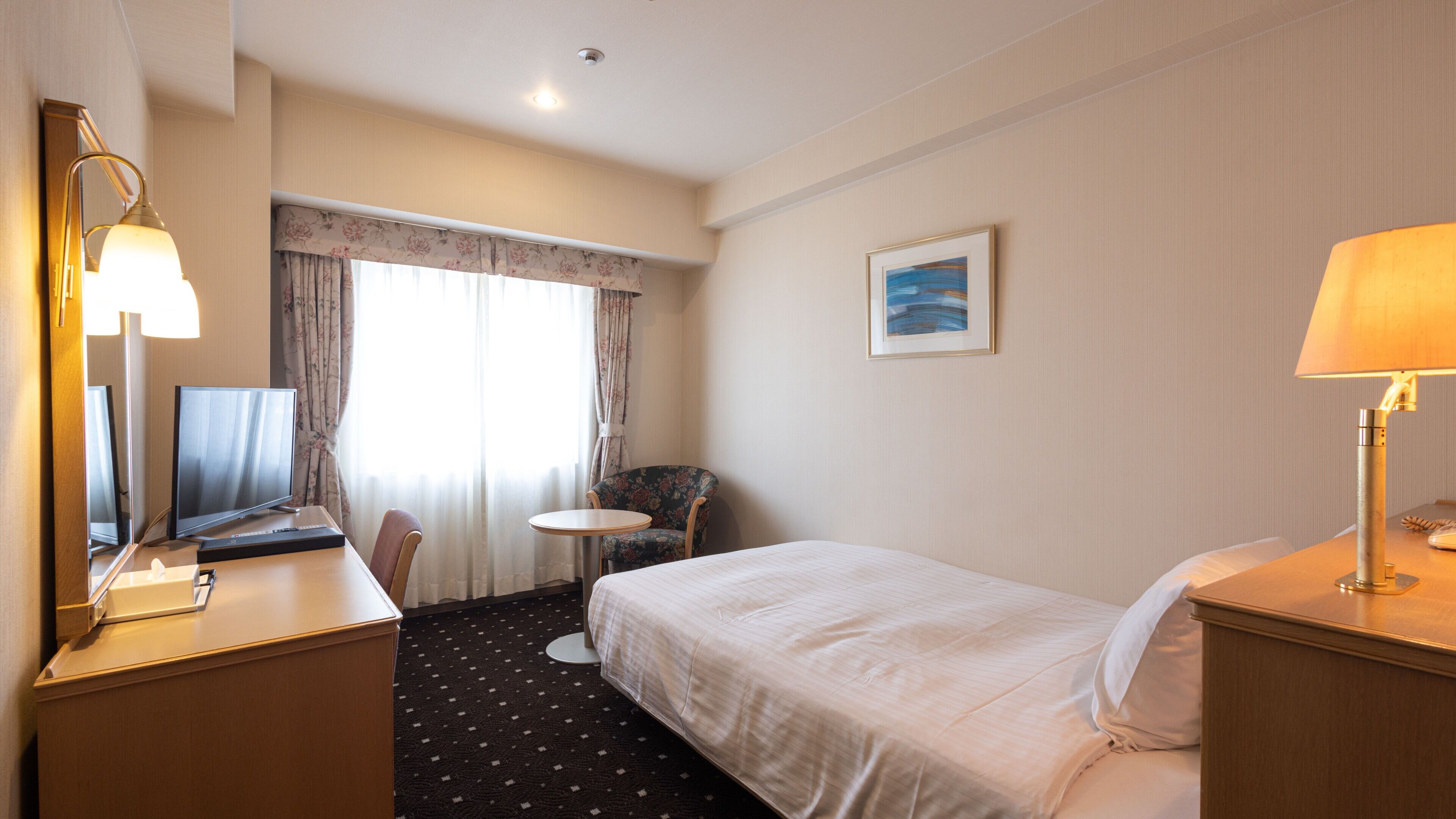 Quarto de hotel com cama, secretária com computador, cadeira, mesinha, abajur, quadro na parede e janela com cortinas.