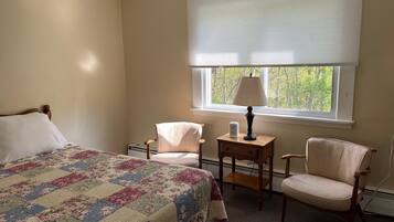 Quarto com cama, duas cadeiras, mesa de cabeceira com abajur e janela com persianas.