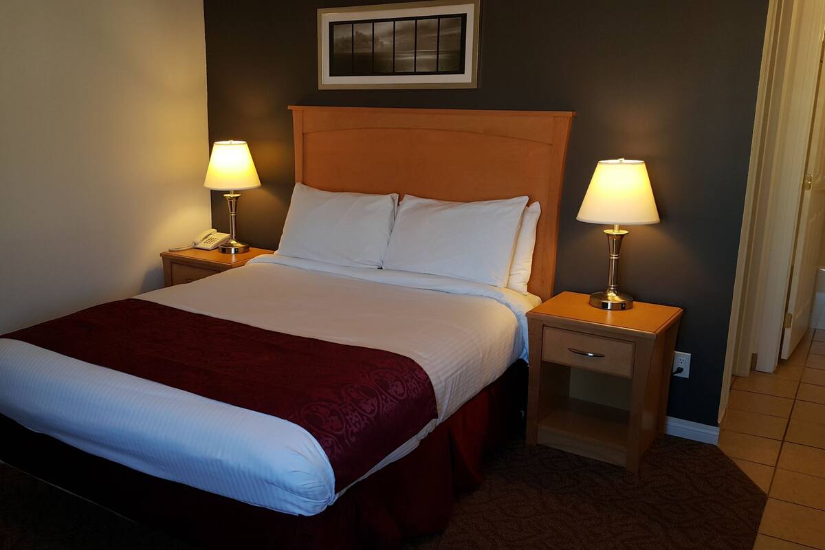 Quarto de hotel com uma cama grande, duas luminárias de cabeceira, um painel de cabeceira de madeira, uma mesa de cabeceira e um quadro na parede.