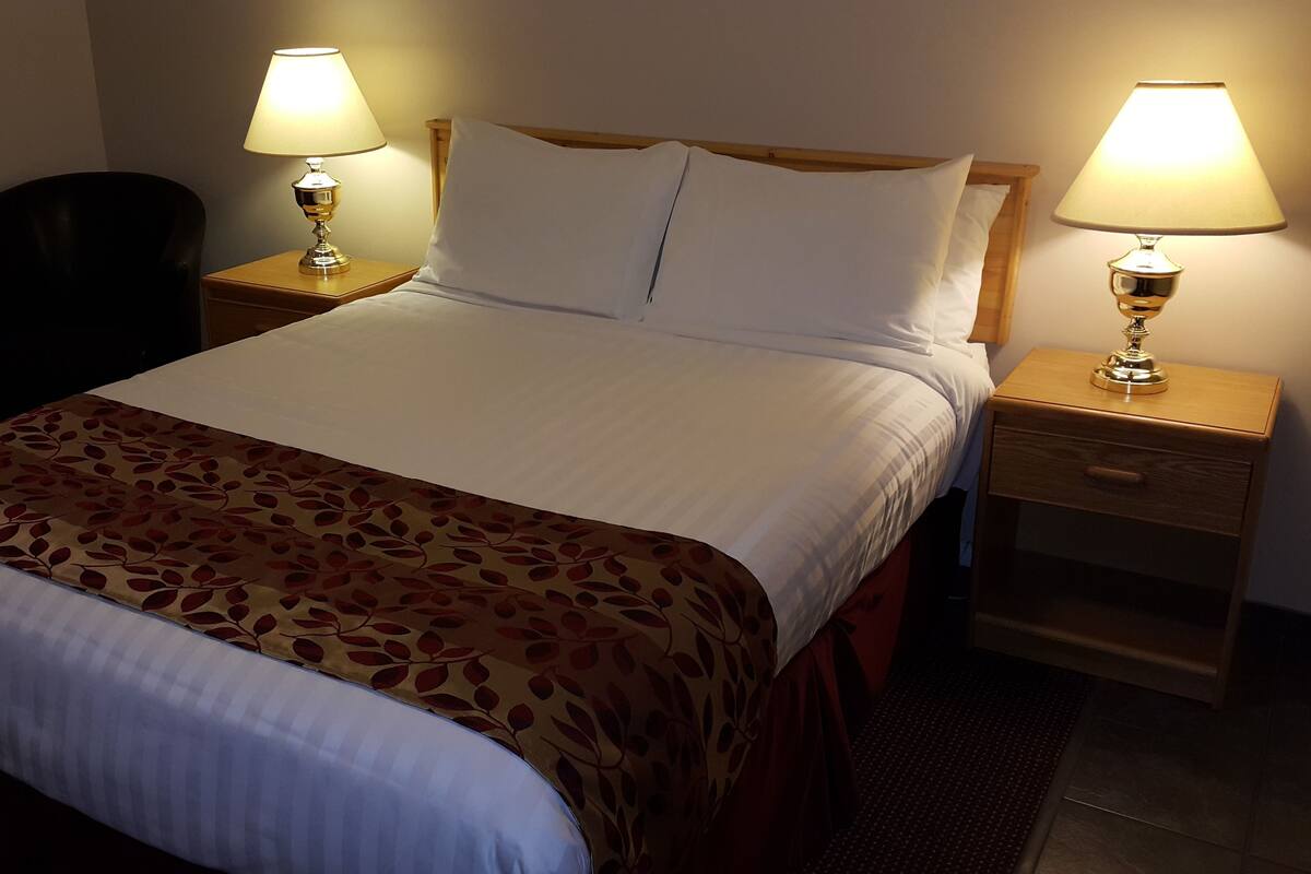Cama bem arrumada com manta estampada, duas luminárias de cabeceira, mesa de cabeceira de madeira e uma cadeira.