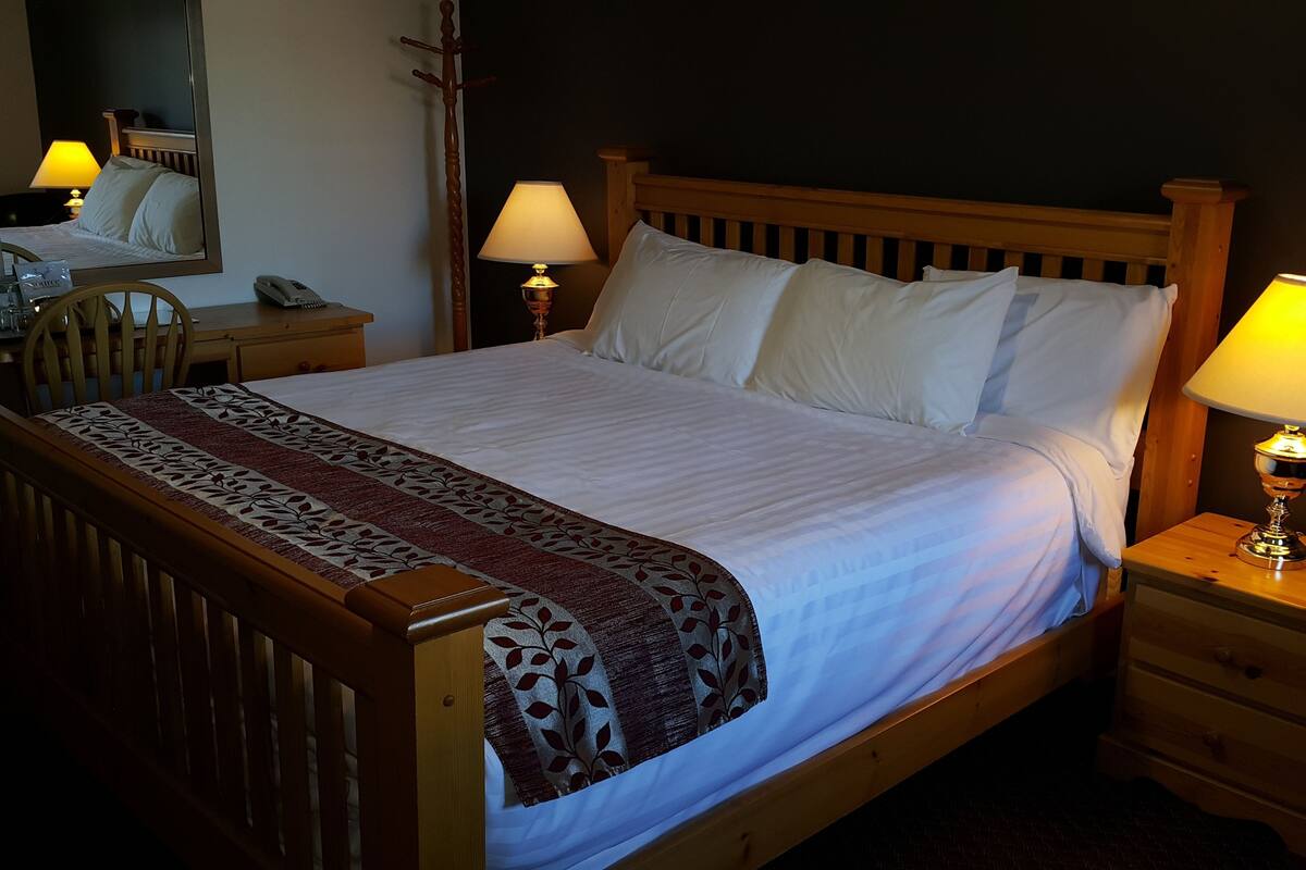 Uma cama bem arrumada com cabeceira de madeira, duas luminárias de cabeceira, uma mesinha de cabeceira e um espelho na parede.