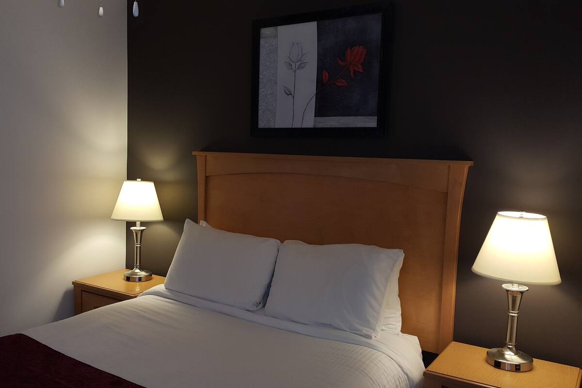 Cama bem arrumada com lençóis brancos, duas luminárias de cabeceira e uma obra de arte emoldurada acima da cabeceira.