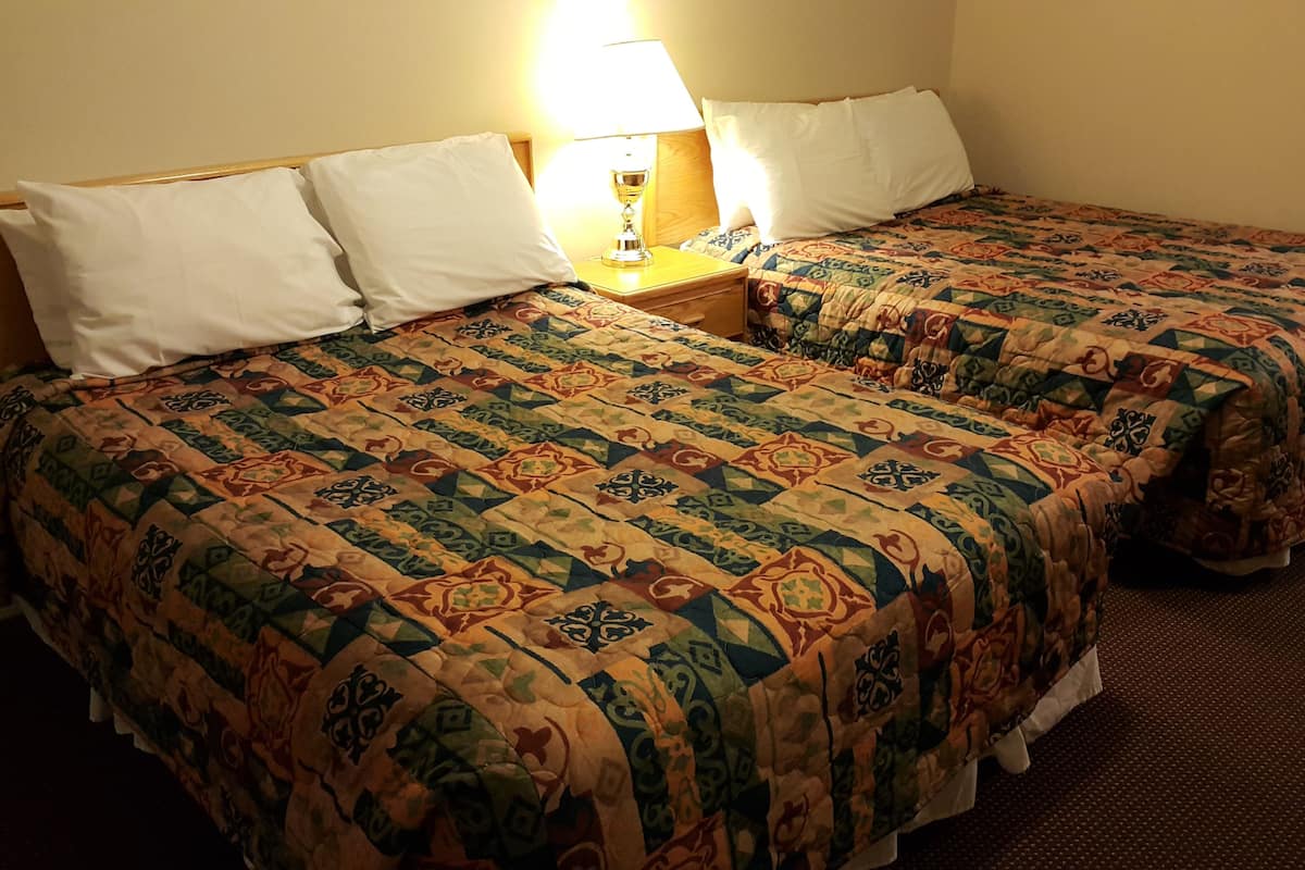 Quarto de hotel com duas camas, cada uma com colcha estampada e almofadas brancas. Há uma mesinha de cabeceira com uma luminária entre as camas.