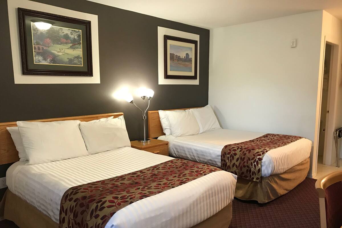 Quarto de hotel com duas camas, uma mesinha de cabeceira com abajur e duas molduras na parede.