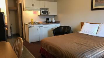 Quarto de hotel compacto com cama, pequeno espaço de cozinha, cadeira e escrivaninha.