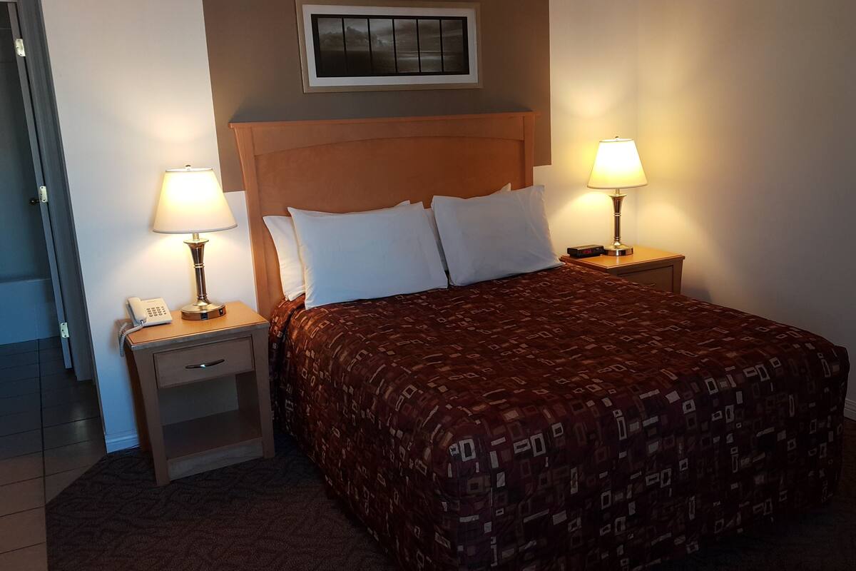 Quarto de hotel com uma cama, duas luminárias de cabeceira, mesa de cabeceira com telefone e janela embutida na parede.