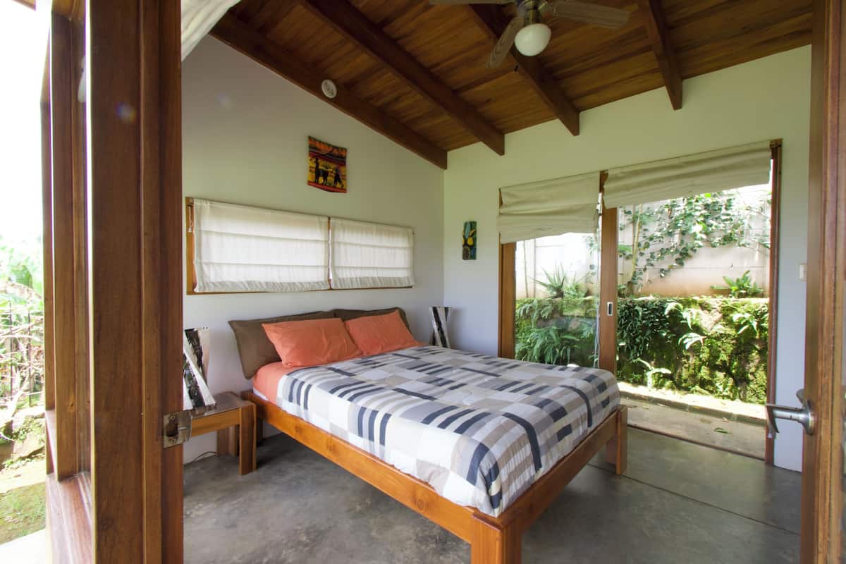 Quarto com cama de madeira, mesa de cabeceira, ventoinha no teto e vista para o verde pela janela.