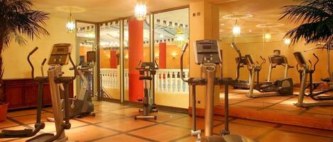 Academia com diversos equipamentos de exercícios, piso de madeira e um espelho grande refletindo os aparelhos.