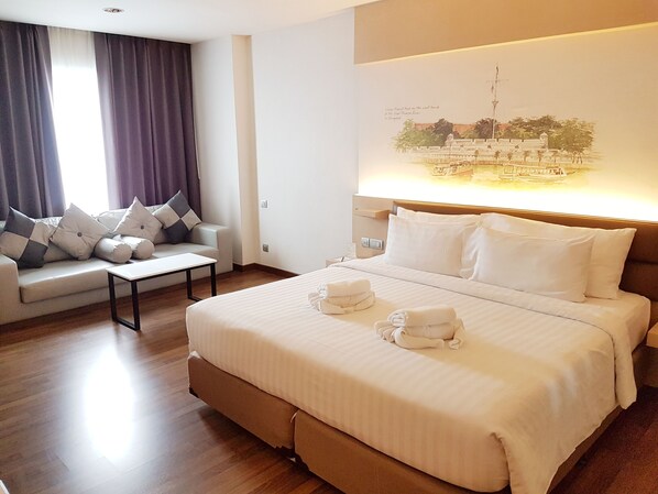 Quarto de hotel com uma cama grande, um sofá com almofadas, uma mesinha e uma luminária na parede.