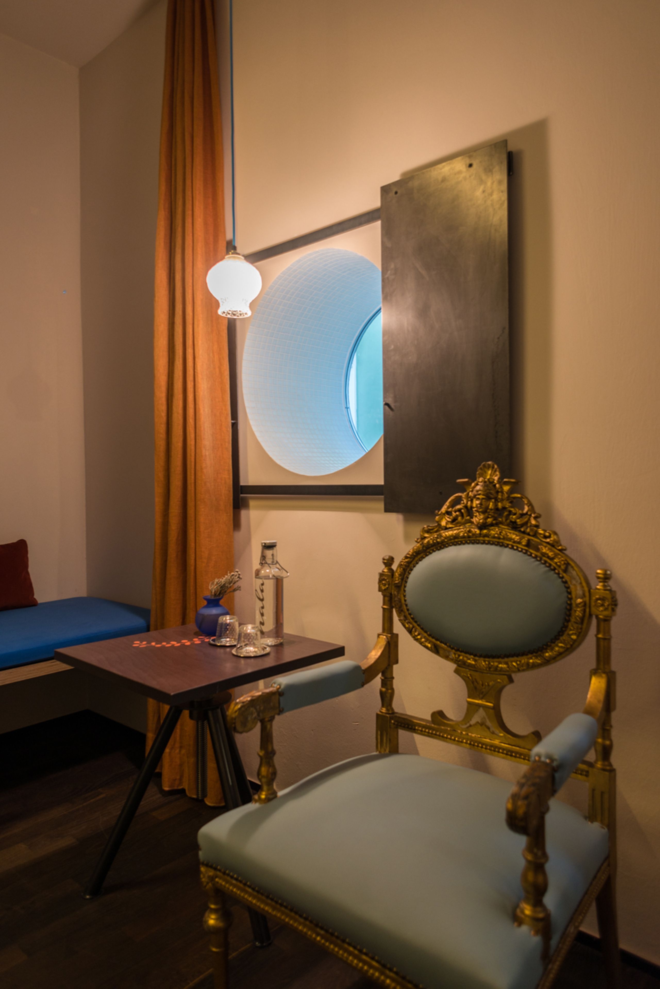 Um quarto com um sofá azul, uma mesa de madeira com uma luminária, um espelho decorativo e uma cadeira com moldura dourada.
