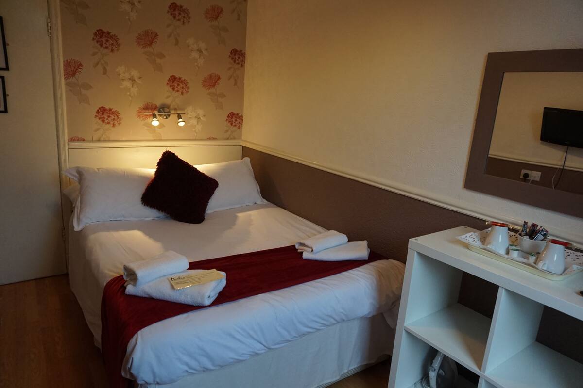 Cama bem arrumada com lençóis brancos e manta vermelha, uma pequena mesa de cabeceira com uma bandeja de xícaras brancas e um controle remoto, e um papel de parede floral na parede.