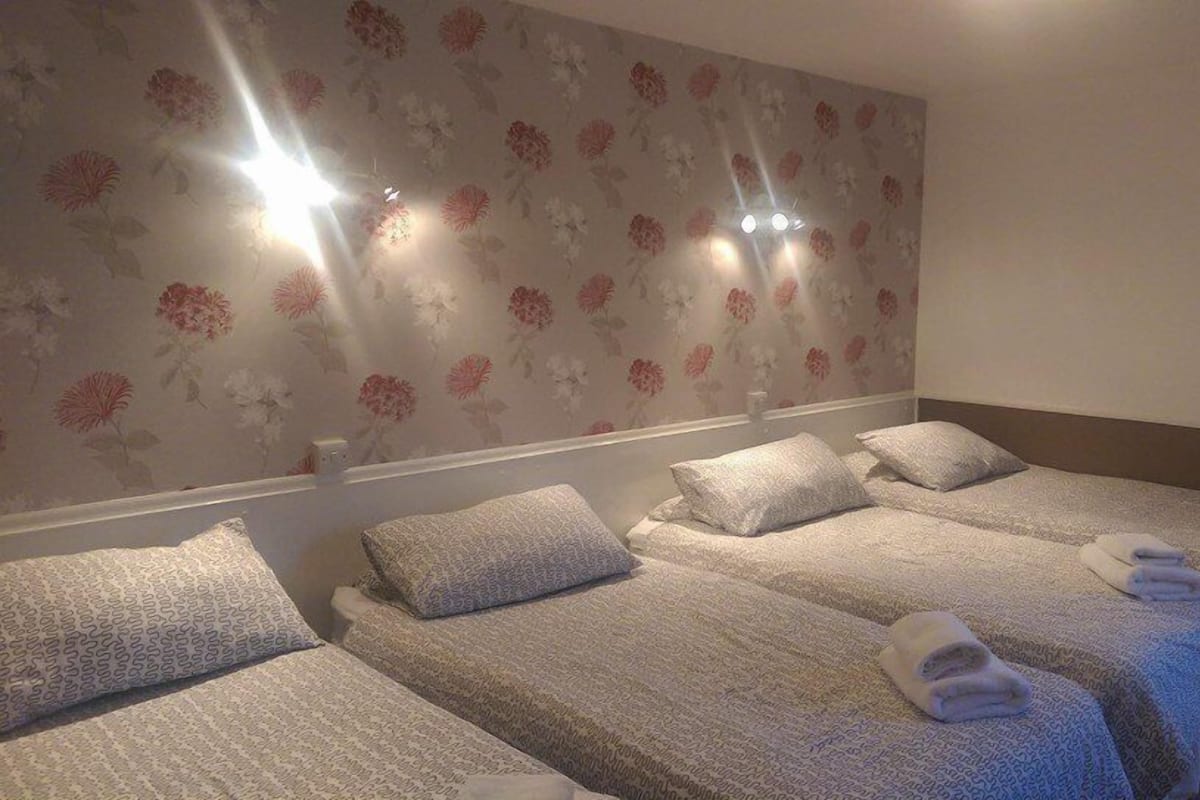 Quarto de hotel com três camas, cada uma com colcha estampada e almofadas. O quarto possui papel de parede floral e duas luminárias fixadas na parede.