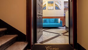 Entrada de hotel com uma escadaria imponente que leva a um sofá azul e uma porta com a inscrição 'Corsio Grand Suite'.