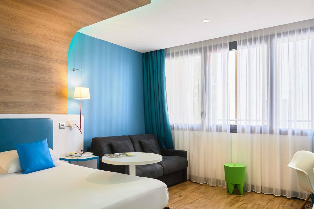 Quarto de hotel com cama, uma mesinha, um sofá, um banquinho verde e janela com cortinas translúcidas.