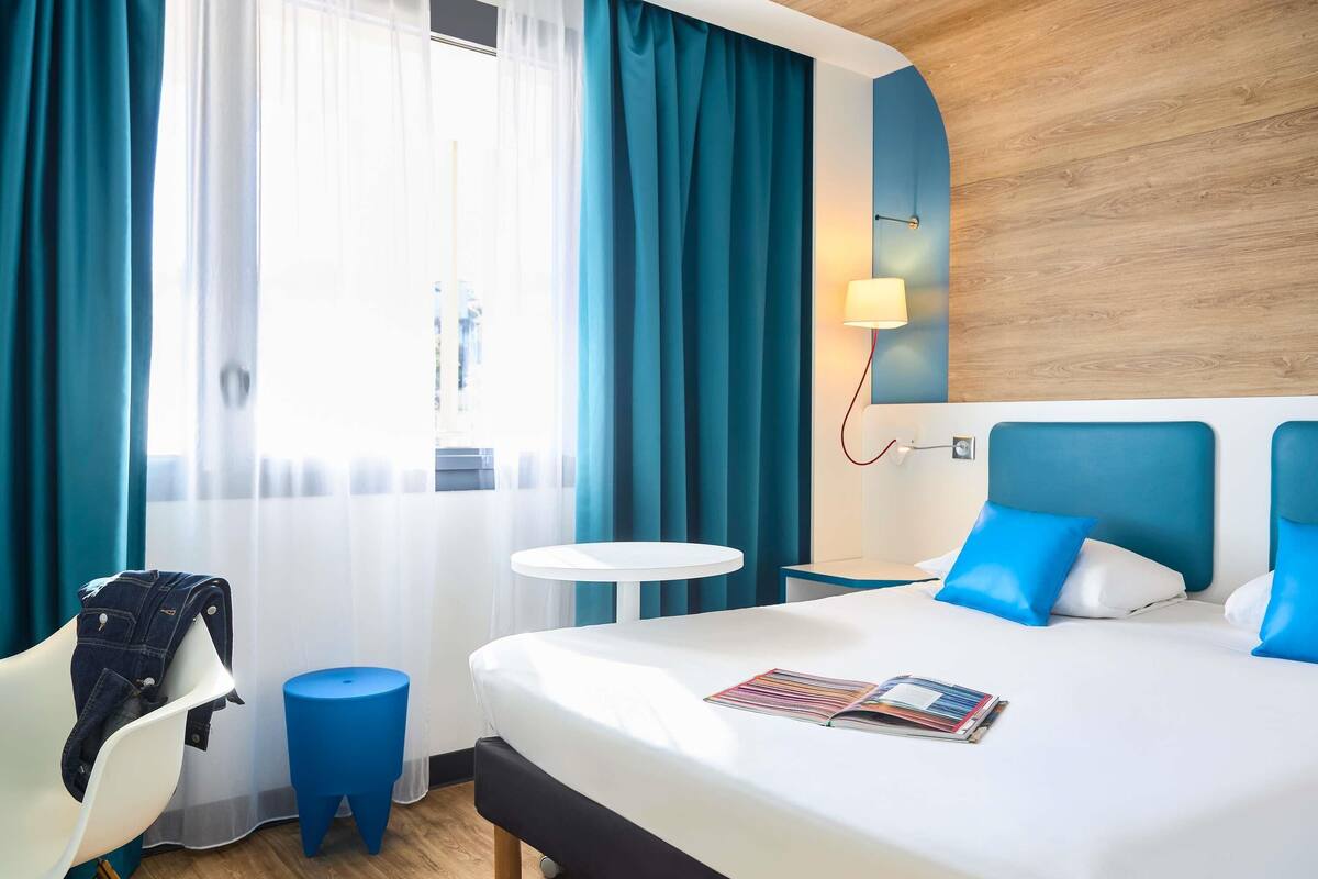 Quarto de hotel com cama, uma mesinha, uma cadeira, luminária e janela com cortinas.