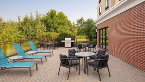 Um pátio externo com cadeiras de lounge e mesas, churrasqueira e um edifício de tijolos ao fundo.
