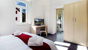 Quarto de hotel moderno com cama, escrivaninha com televisão, guarda-roupa e janela com vista para árvores.