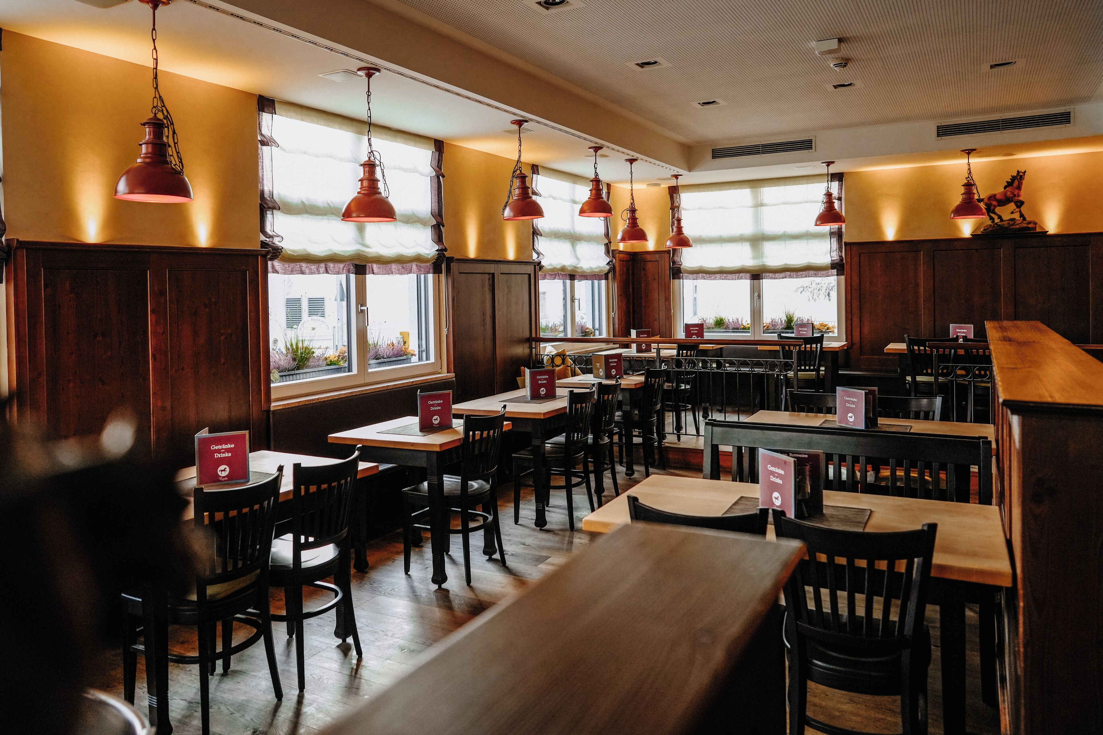 Restaurante com mesas e cadeiras de madeira, luminárias pendentes vermelhas e grandes janelas com cortinas.