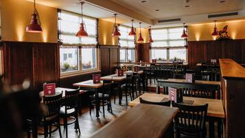 Restaurante com mesas e cadeiras de madeira, luminárias pendentes vermelhas e grandes janelas com cortinas.