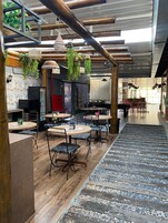 Interior de restaurante com mesas de madeira, plantas pendentes e parede revestida de azulejos.
