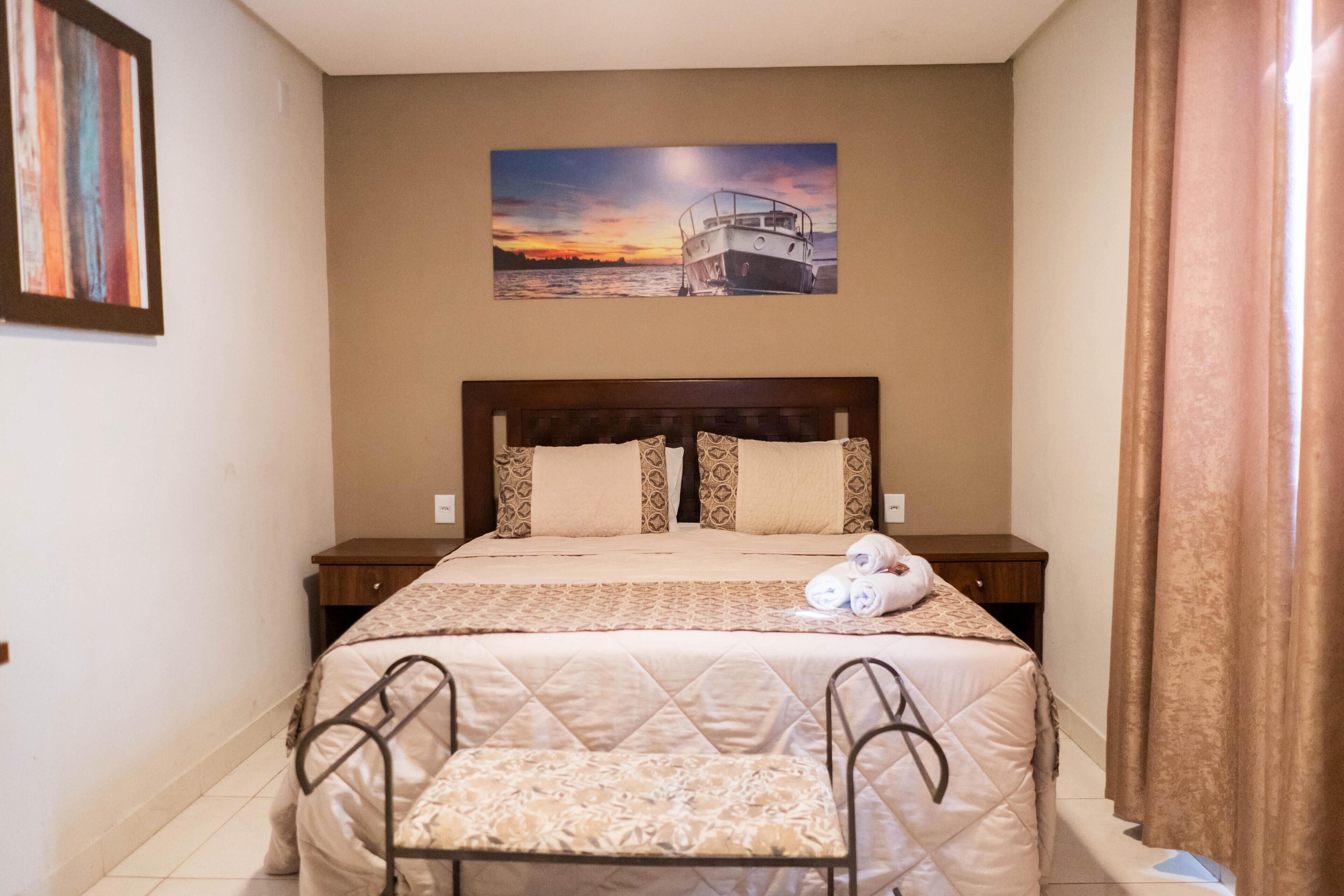 Quarto de hotel com cama, duas mesas de cabeceira, uma cadeira, um quadro de um barco na parede e uma cortina.