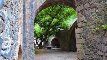 Um corredor de pedra arqueado com uma árvore no centro, que leva a outra estrutura arqueada ao longe.