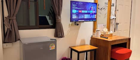 Quarto com televisão fixada na parede exibindo a interface de um serviço de streaming, uma pequena mesa de madeira, uma cadeira vermelha, uma chaleira branca e uma lixeira.