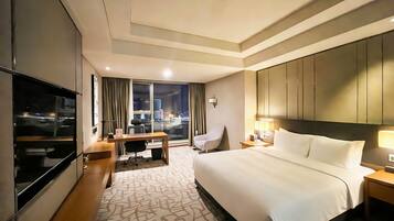 Quarto de hotel com uma cama grande, uma escrivaninha, uma cadeira, uma televisão e vista para a cidade à noite.