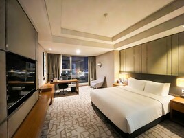 Quarto de hotel com uma cama grande, uma escrivaninha, uma cadeira, uma televisão e vista para a cidade à noite.