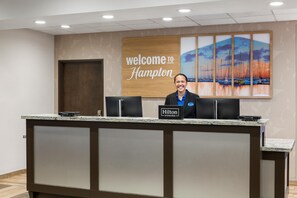Balcão de recepção de hotel com um letreiro que diz 'Welcome to Hampton', uma placa de identificação 'Hilton Honors' e um painel com barcos à vela ao fundo.