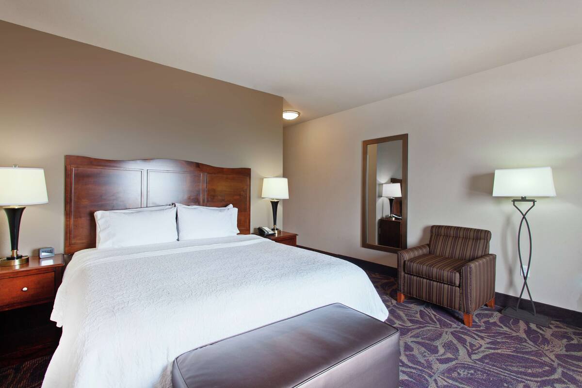 Quarto de hotel com uma cama grande, uma cadeira, mesa de cabeceira, abajur e espelho.