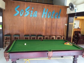 Uma mesa de bilhar com superfície de feltro verde, uma bola branca e um conjunto de bolas de bilhar. Seis cadeiras de madeira estão alinhadas contra uma parede de madeira com o texto "Sosia Hotel" em letras azuis.