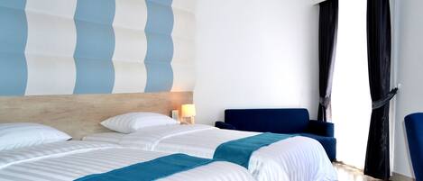 Quarto de hotel com duas camas, cabeceira de madeira, sofá azul e uma janela grande com cortinas pretas.