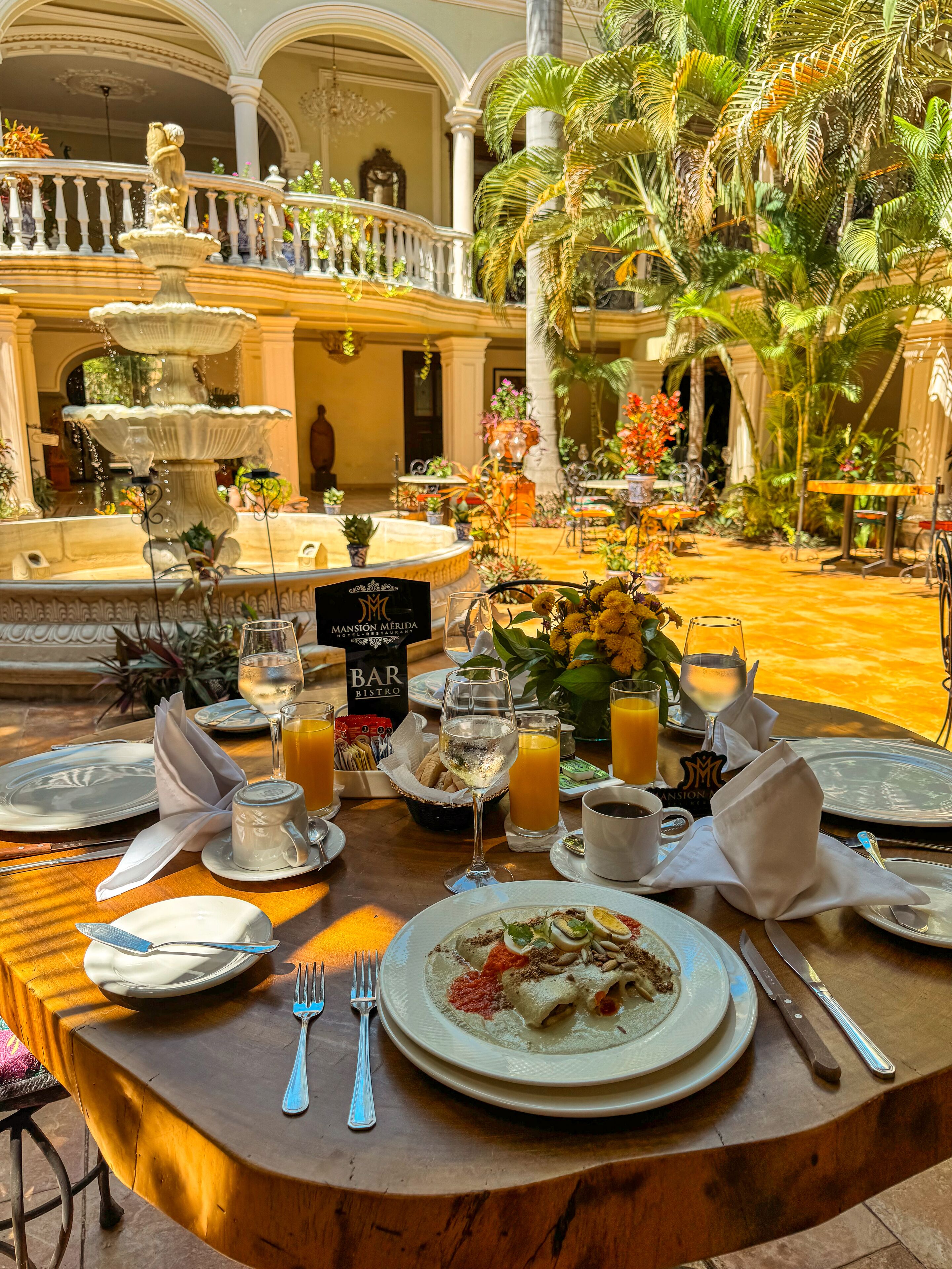 Mesa de jantar posta para o café da manhã, com vista para uma fonte e plantas tropicais.