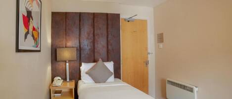 Quarto de hotel com uma cama, mesa de cabeceira com abajur, telefone, radiador e porta.