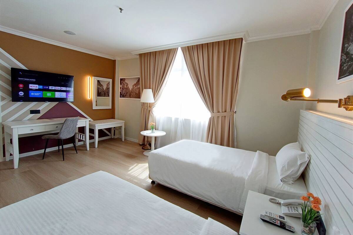 Quarto de hotel com uma cama, uma escrivaninha com cadeira, televisão fixada na parede e abajur numa mesinha de cabeceira.
