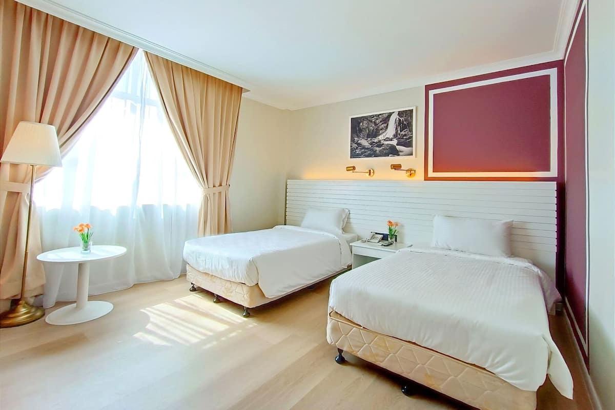Quarto de hotel com duas camas, uma mesa de cabeceira com abajur, uma mesinha redonda com flores e um quadro na parede.