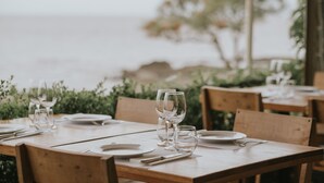 Mesa de jantar de madeira posta com pratos, copos e talheres, com vista para um corpo d'água.
