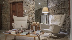 Um quarto aconchegante com parede de pedra, porta de madeira, um espelho grande e duas cadeiras. Há uma mesa com livros e uma luminária pequena.