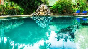 Piscina translúcida com uma estrutura de pedra no centro, rodeada por vegetação exuberante e espreguiçadeiras azuis.