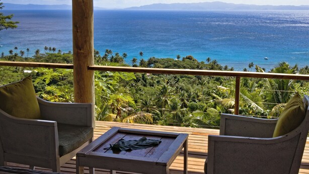 Um deck de madeira com duas cadeiras e uma mesa, com vista para uma paisagem tropical com palmeiras e o oceano.