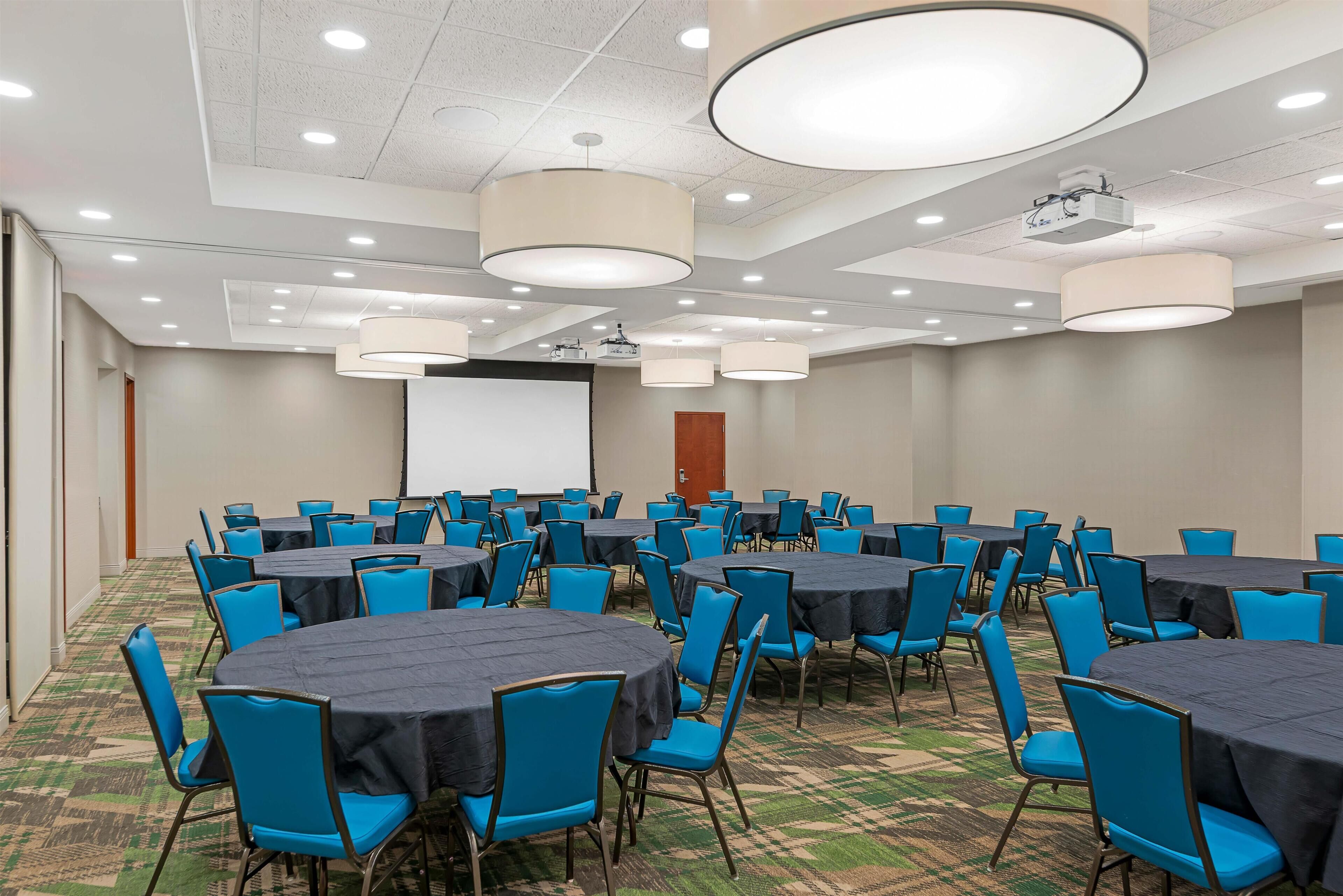 Sala de conferências com várias mesas redondas e cadeiras azuis, equipada com projetores instalados no teto e iluminação ambiente.