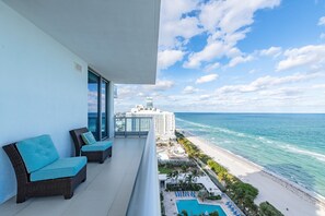 Varanda com mobiliário de vime com vista para uma praia e um hotel ao fundo.