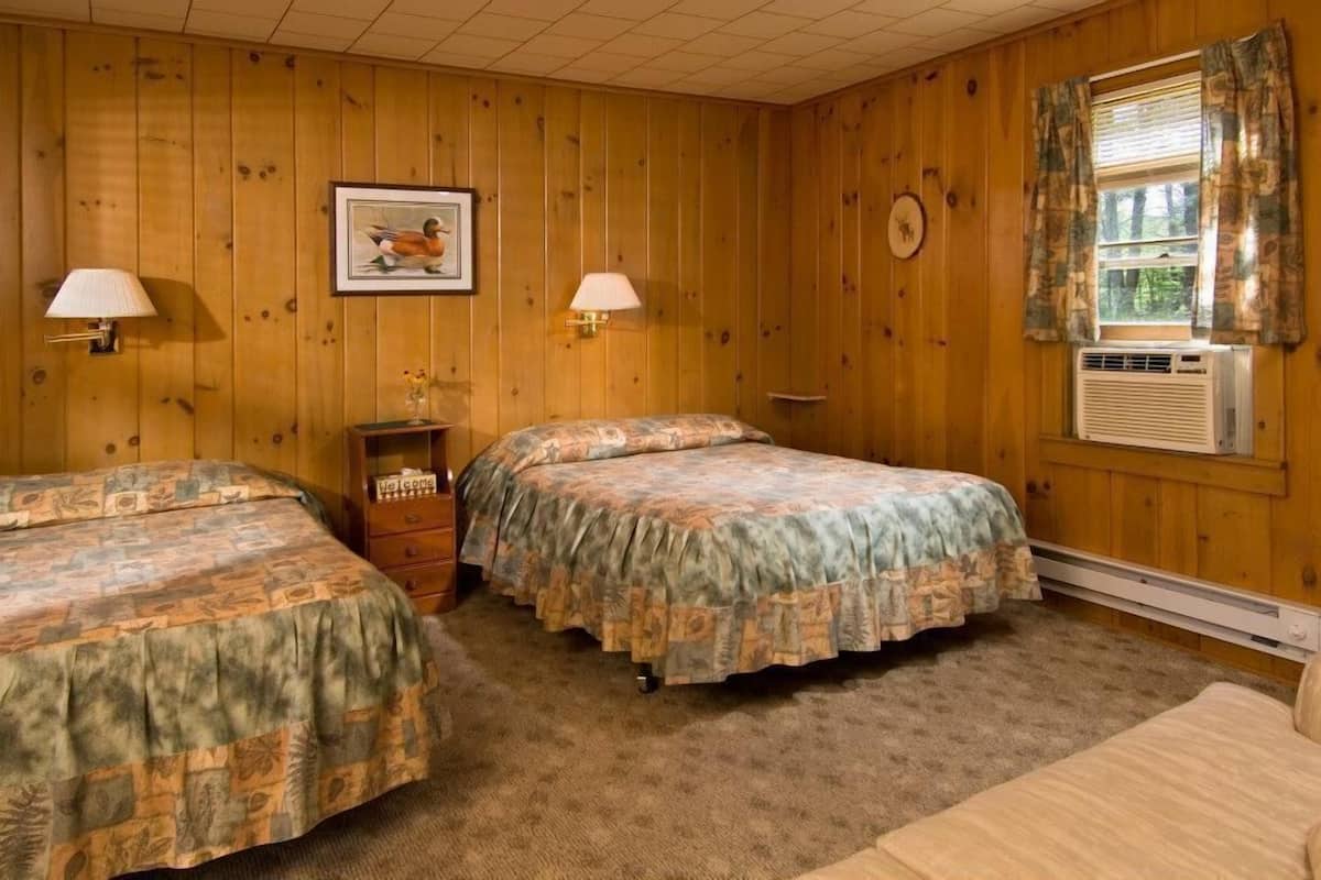 Quarto com duas camas, paredes de madeira, um quadro emoldurado, duas luminárias de parede, uma janela com cortinas e um aparelho de ar condicionado.
