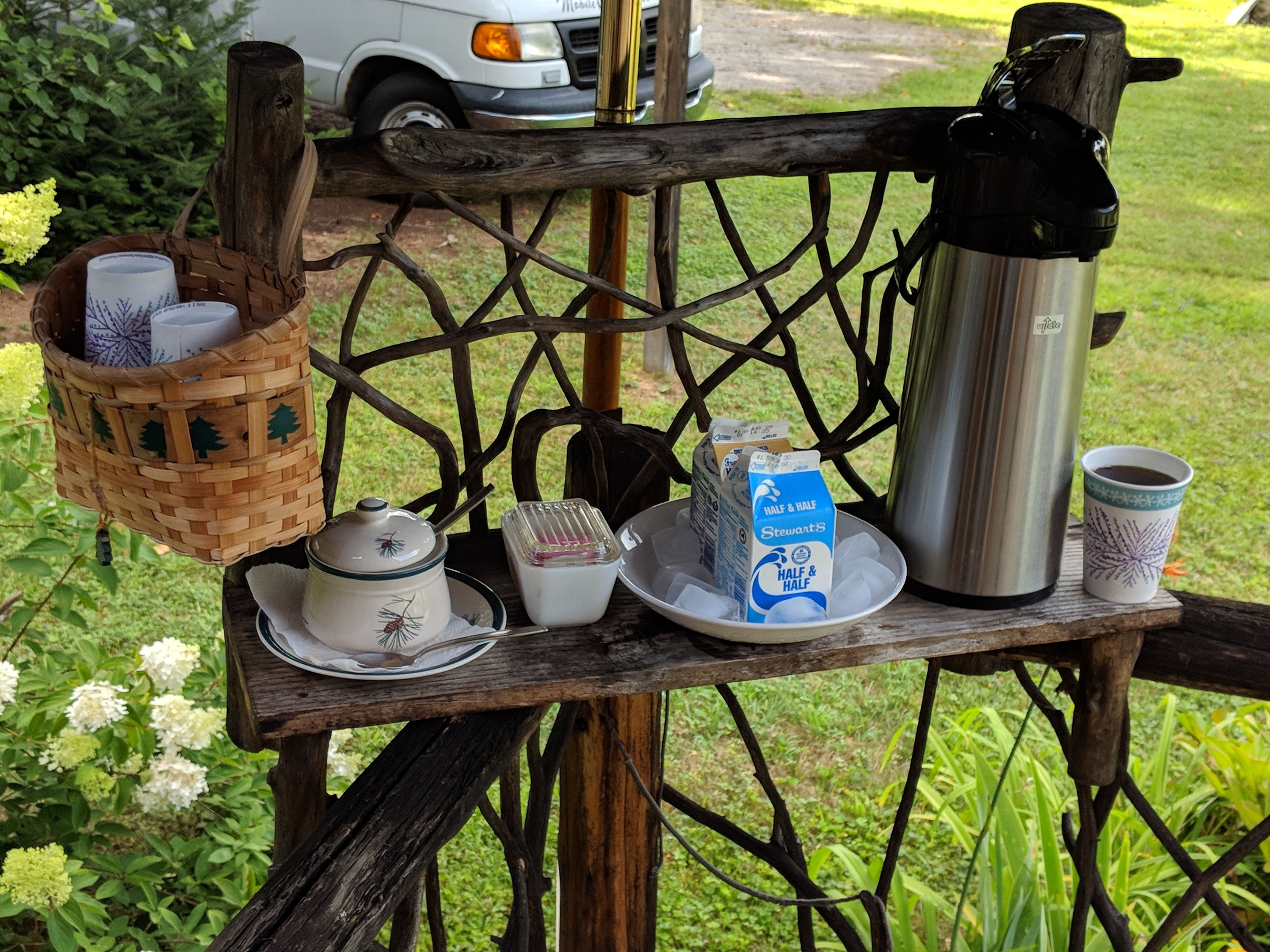 Um café da manhã ao ar livre rústico, com uma cesta de vime, uma garrafa térmica, um recipiente com leite, um bule e uma xícara sobre uma mesa de madeira.