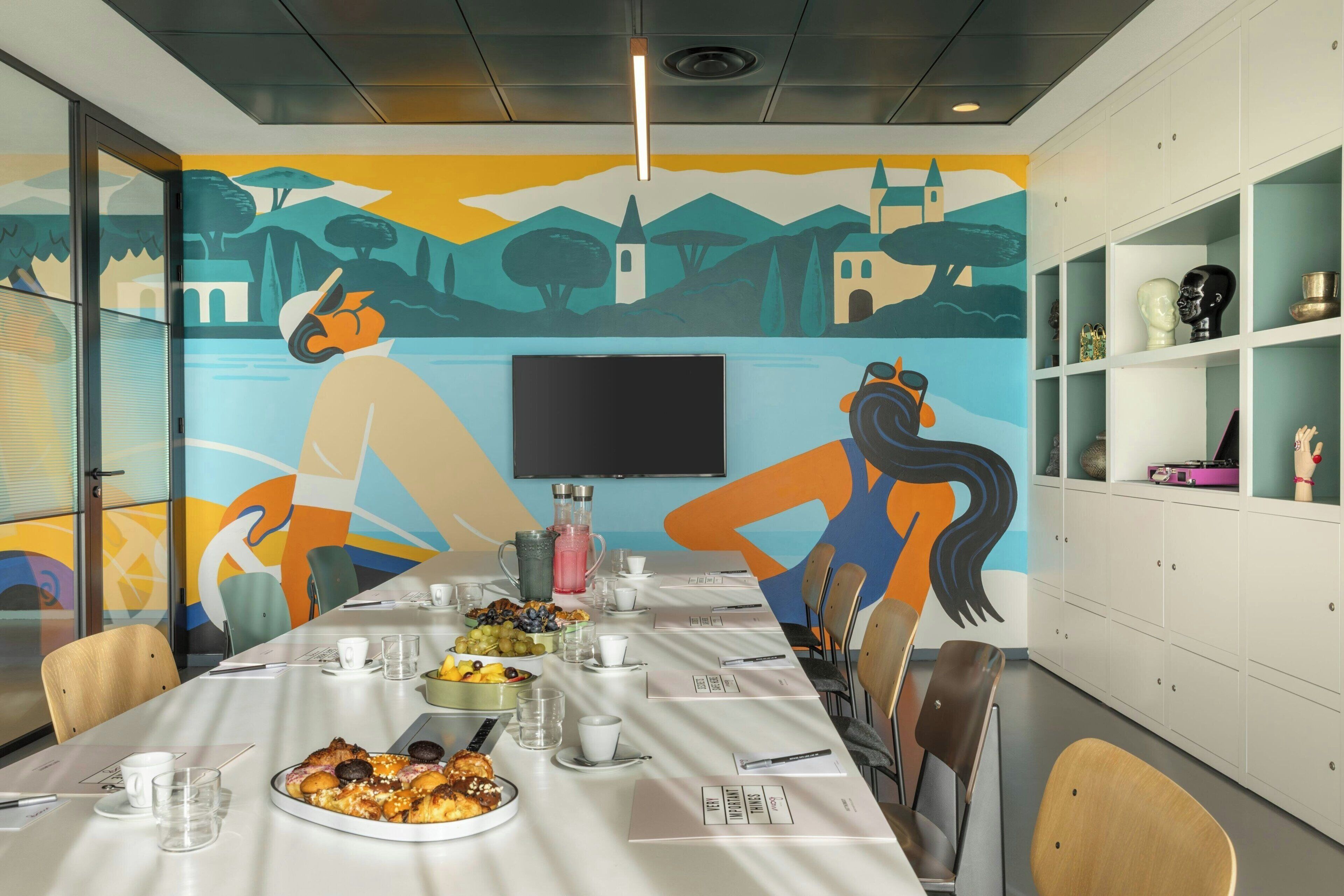 Uma sala de conferências com uma mesa comprida preparada para uma reunião, um mural na parede e uma estante branca com objetos decorativos.