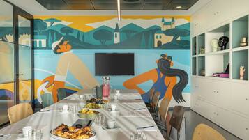 Uma sala de conferências com uma mesa comprida preparada para uma reunião, um mural na parede e uma estante branca com objetos decorativos.
