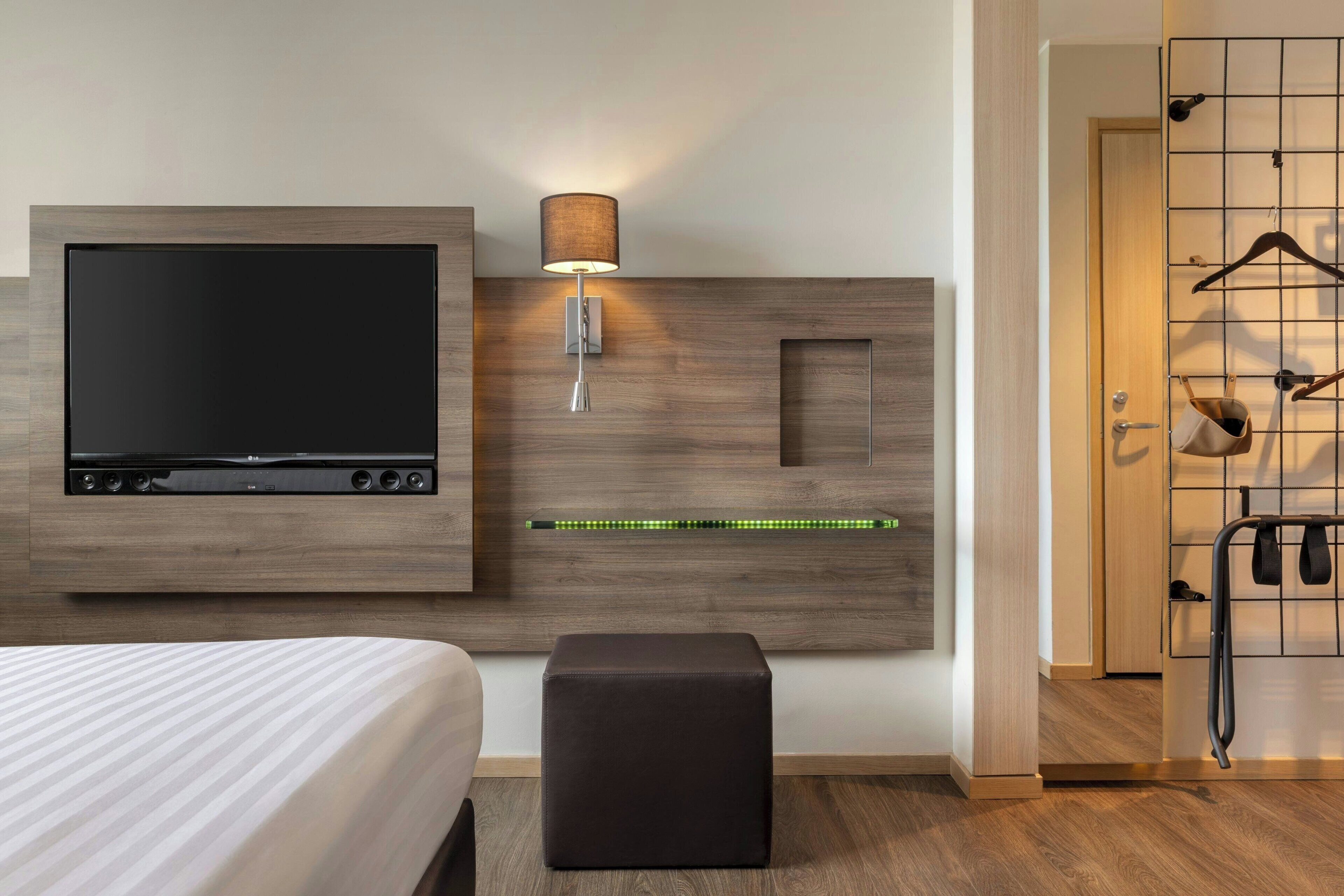 Quarto de hotel moderno com televisão de tela plana, luminária de parede e sapateira.